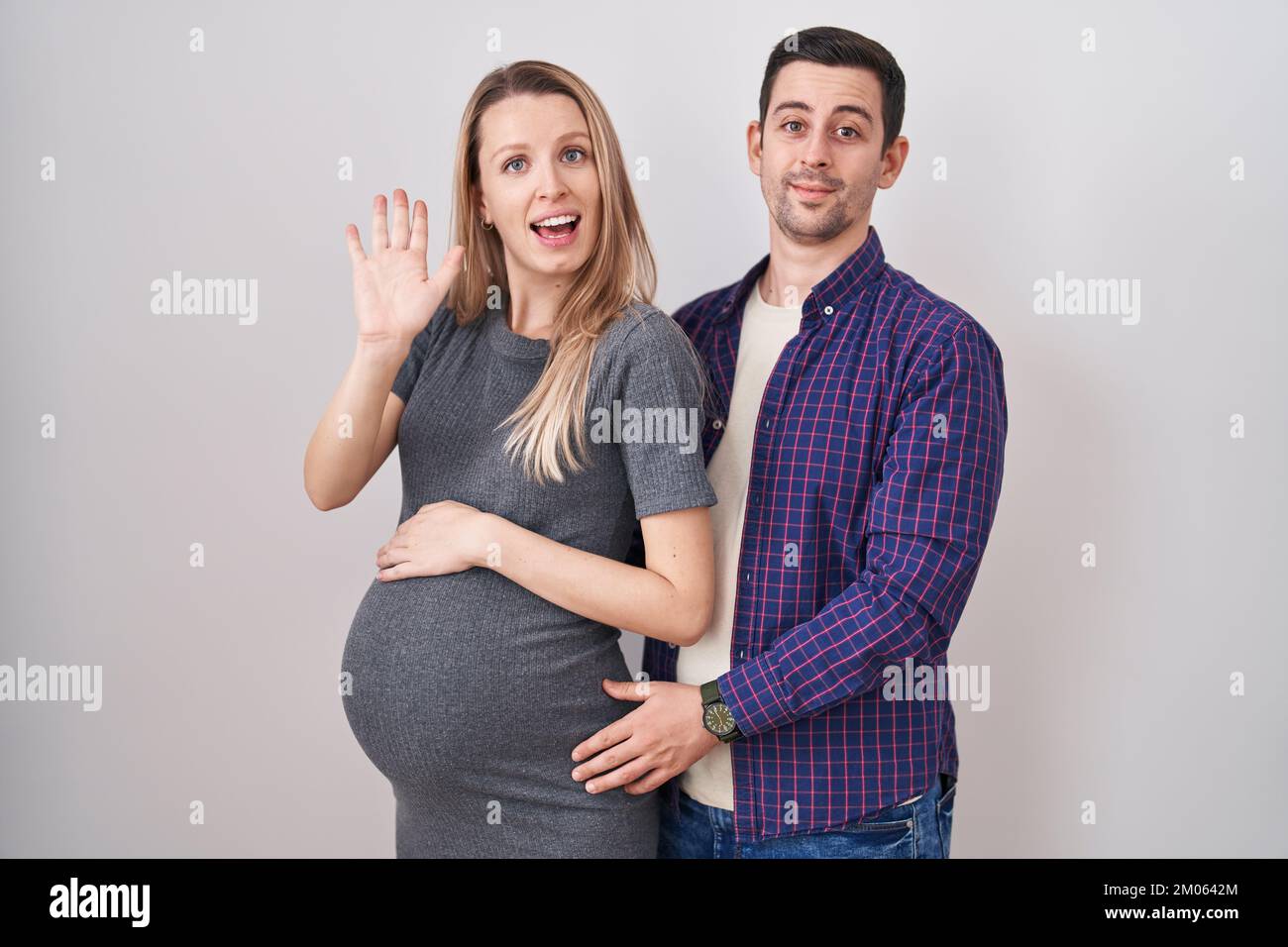 Young couple expecting a baby standing over white background waiving ...