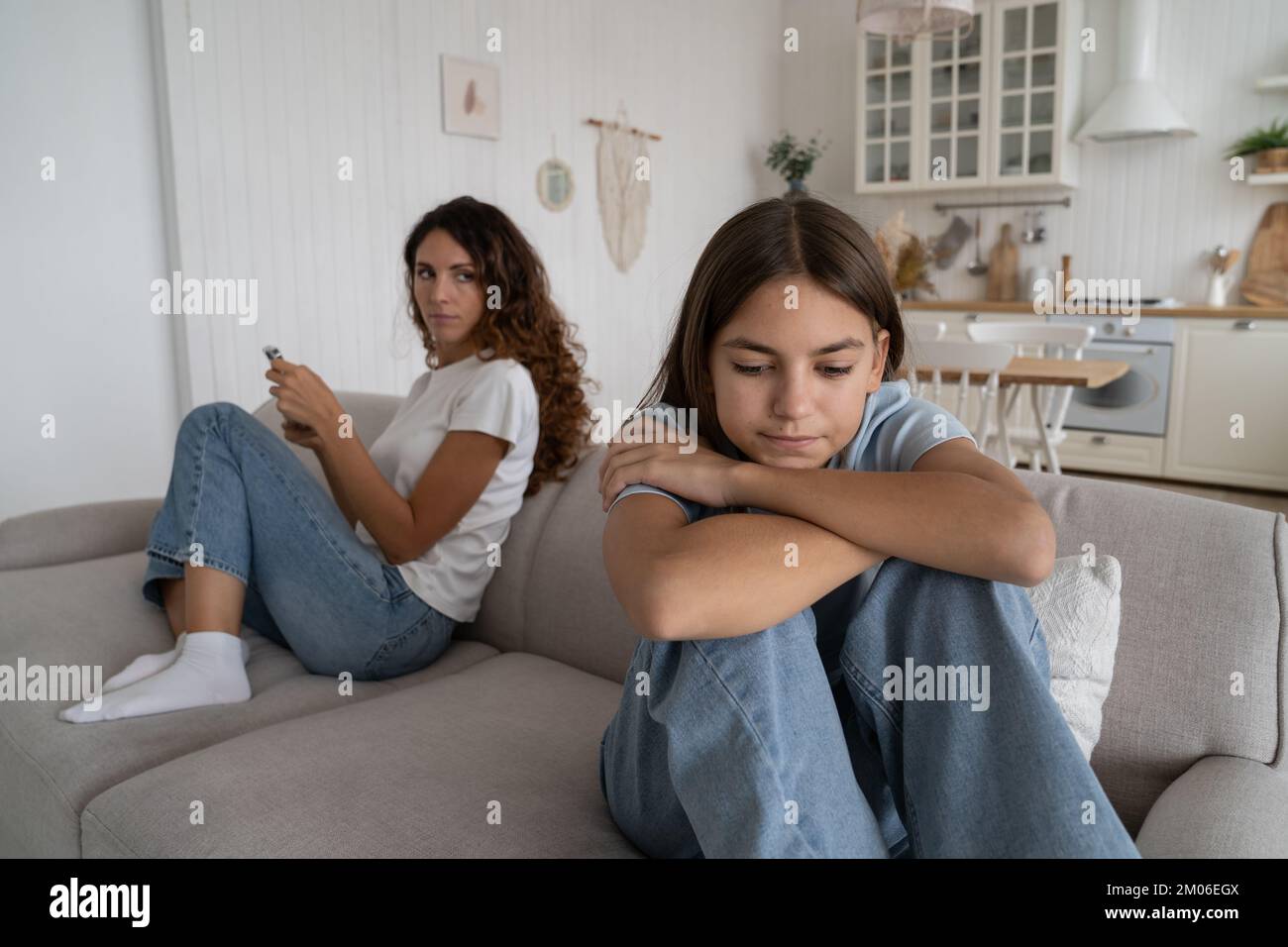 Casual worried woman sits on sofa behind sad teen girl leans on knees ...