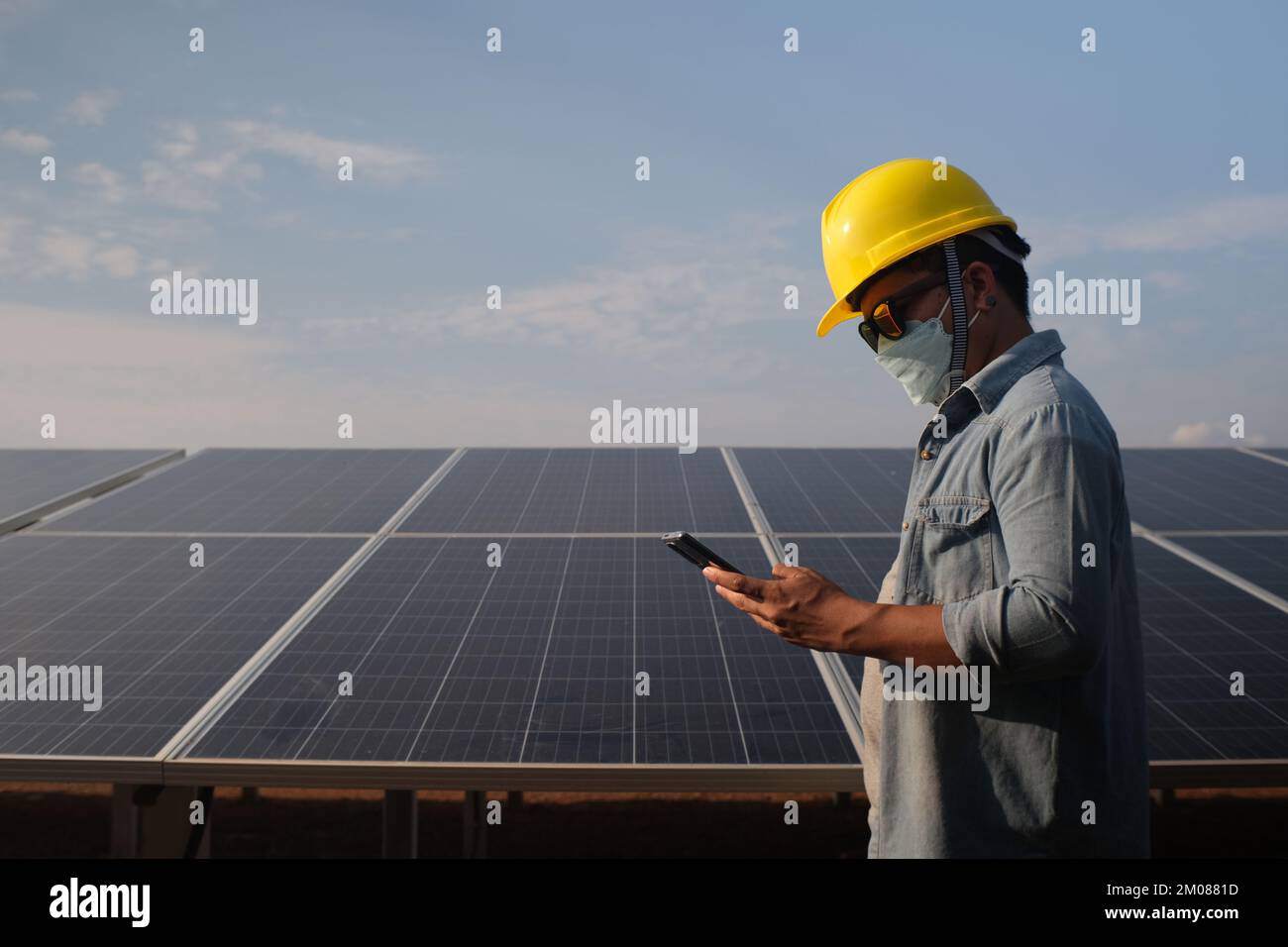 A contractor wearing a yellow helmet and mask is pressing a mobile phone against a backdrop of ...
