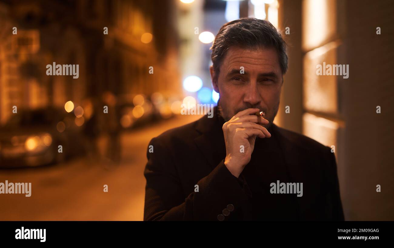 Mid adult man smoking cigar on the street at night wearing black coat ...