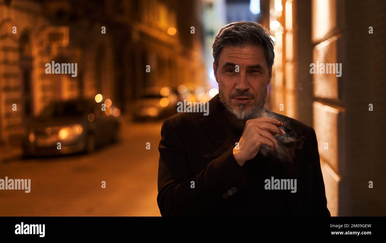 Mid adult man smoking cigar on the street at night wearing black coat ...