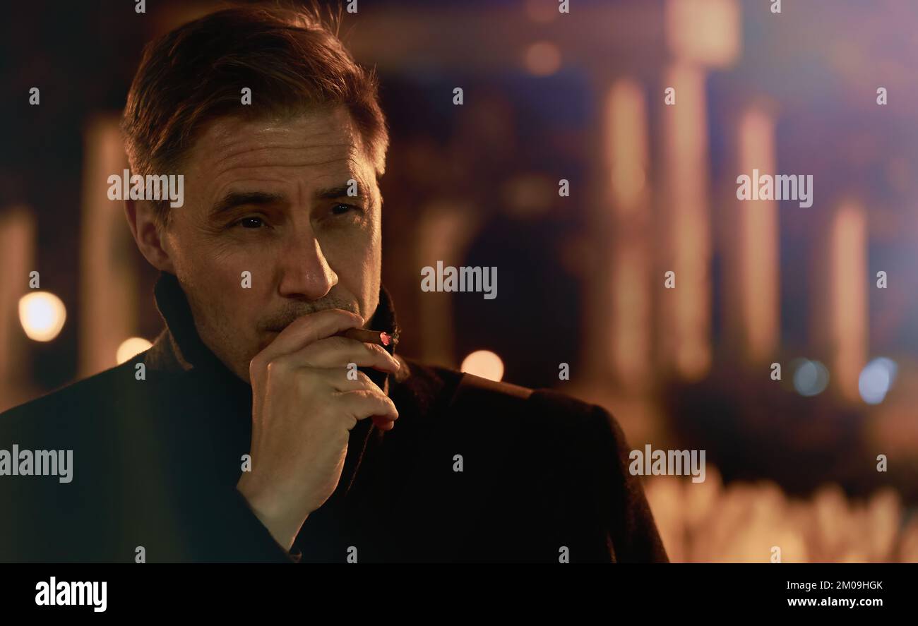 Mid adult man smoking cigar on the street at night wearing black coat ...