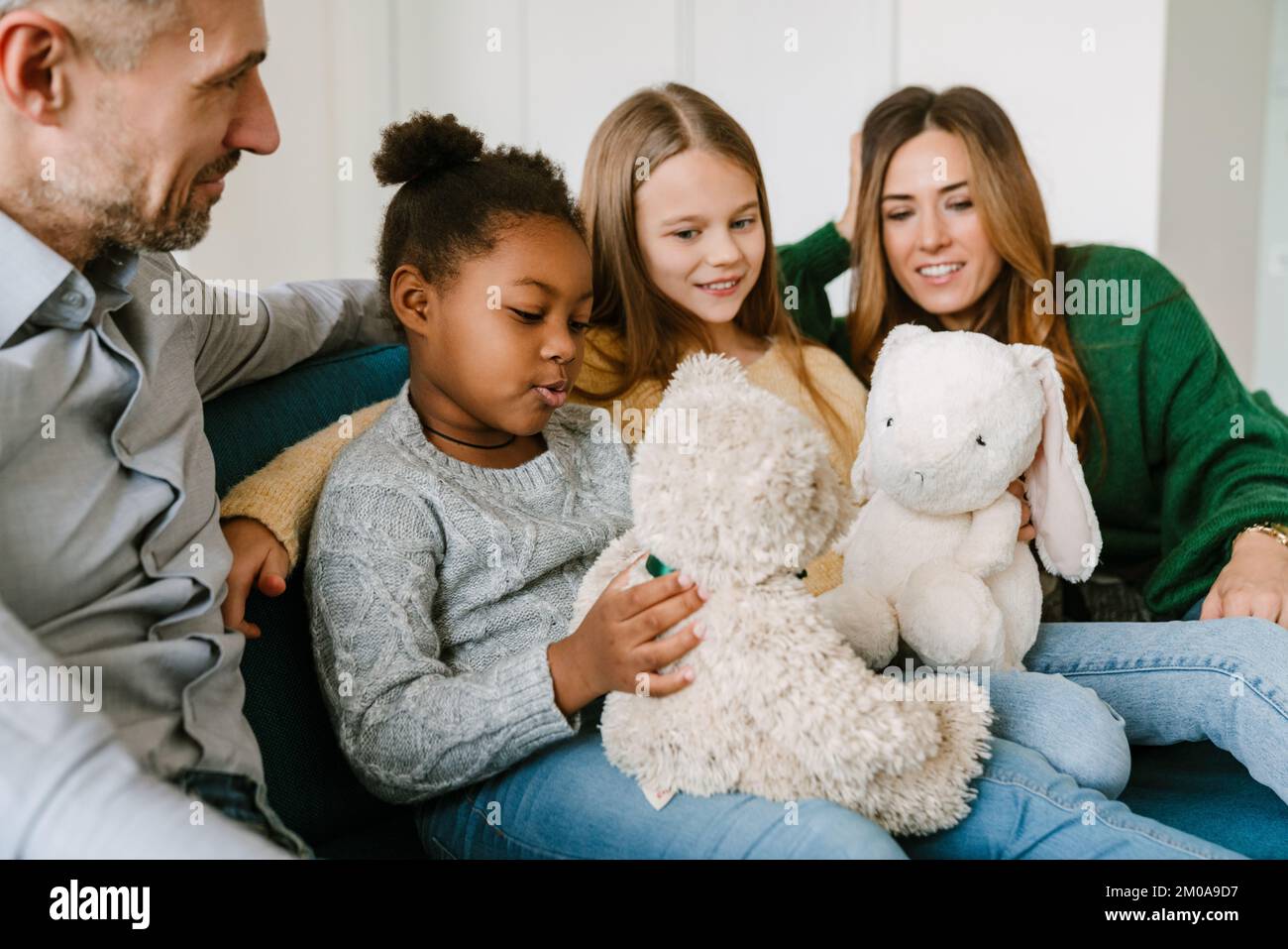 Happy family sitting on couch, adopted sisters playing stuffed toys ...