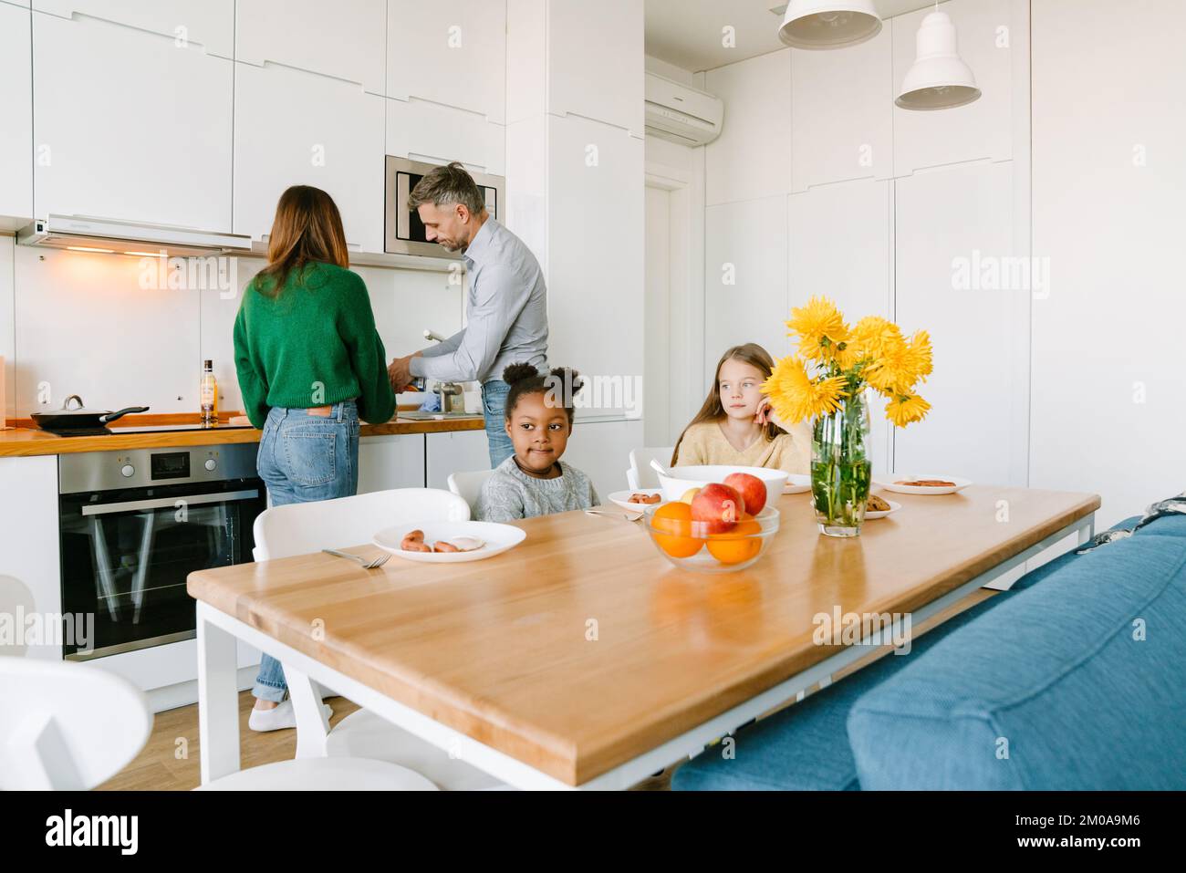 Adopted sisters waiting for breakfast near parents in cozy kitchen at ...