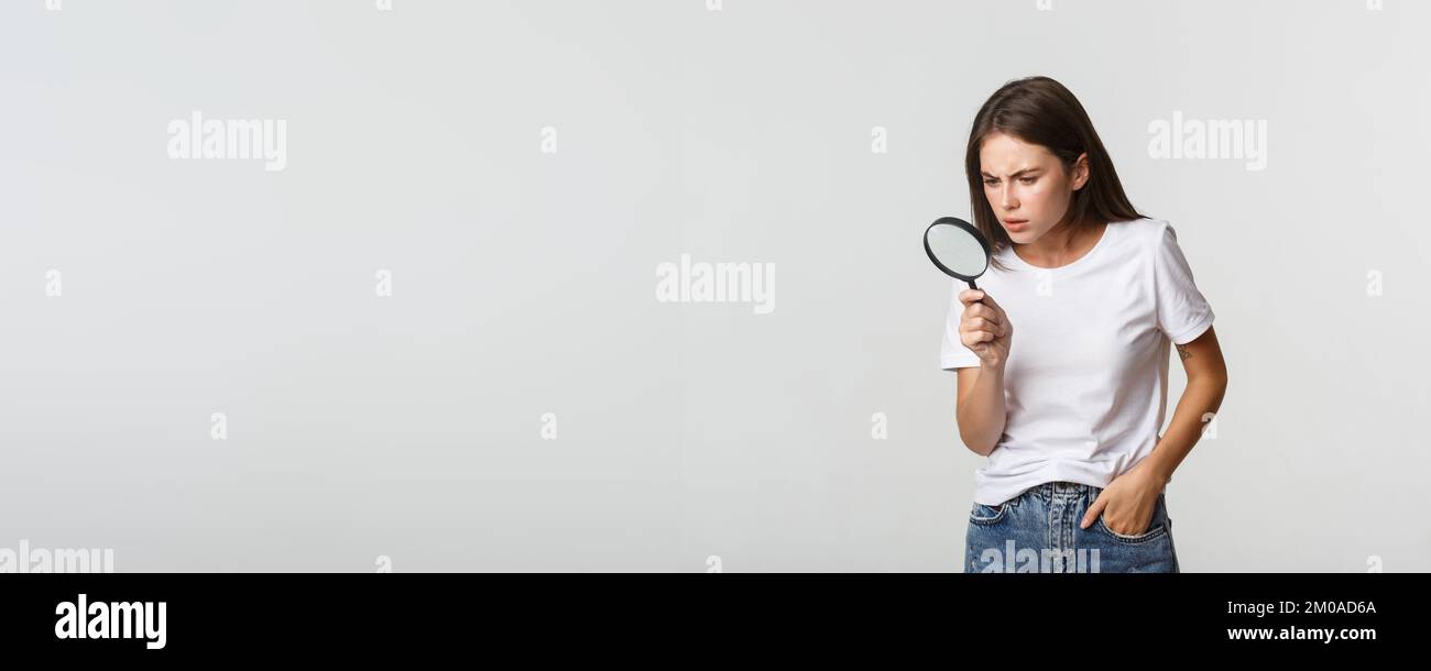 Serious-looking smart beautiful girl looking through magnifying glass ...