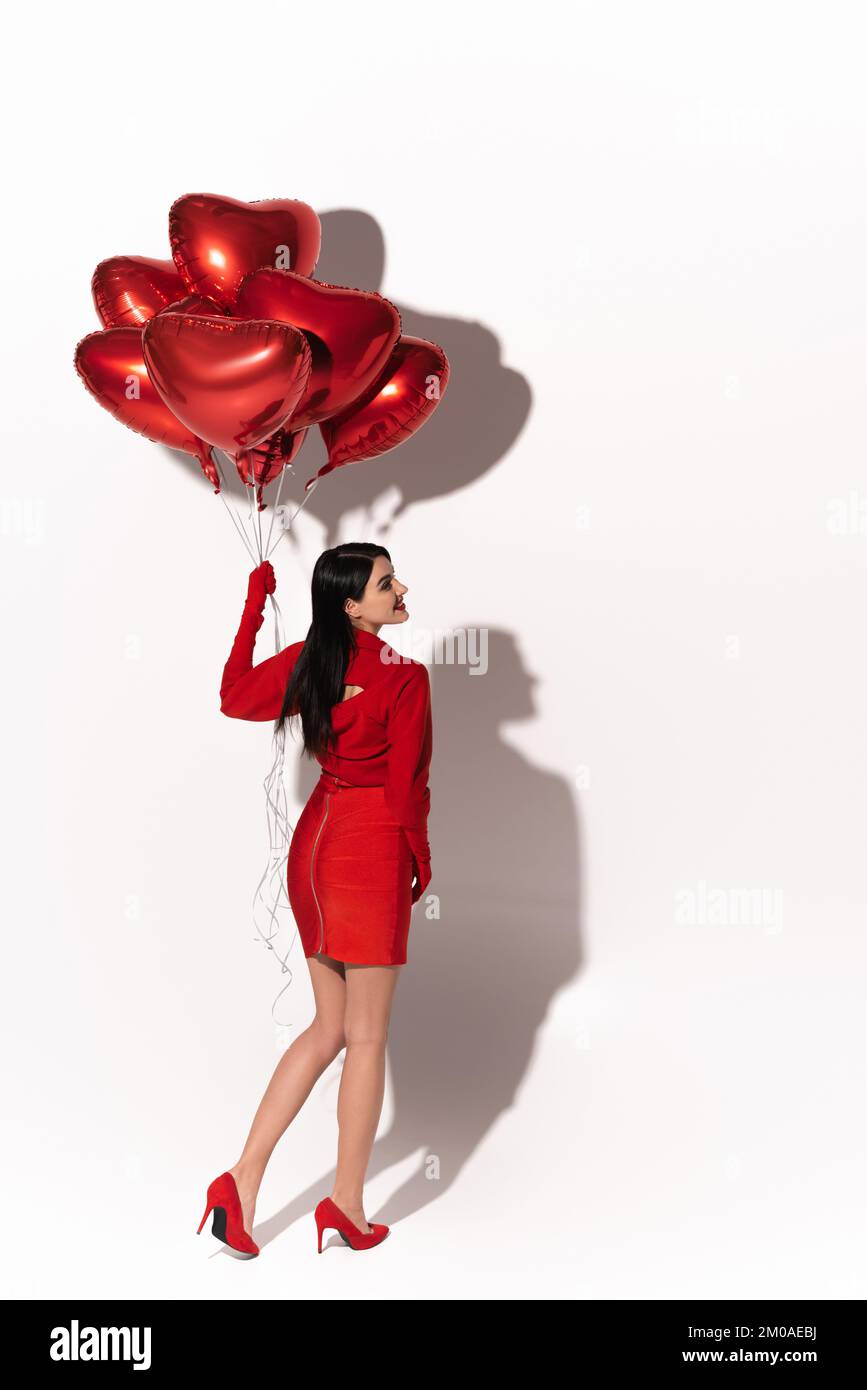 Side view of smiling model in red dress and heels holding festive heart ...