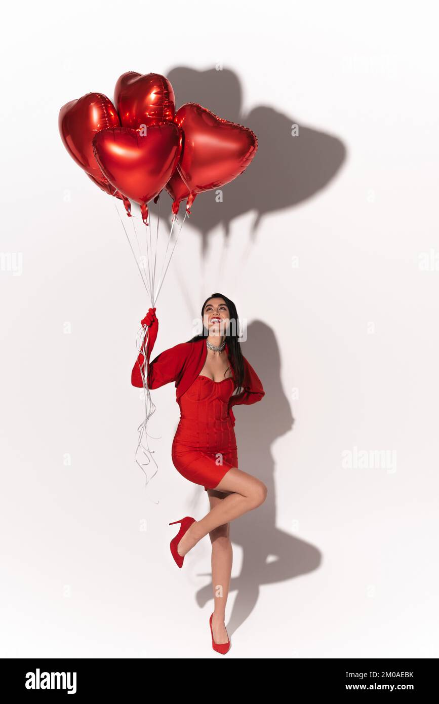 Stylish woman in red dress and heels looking at balloons in heart shape ...