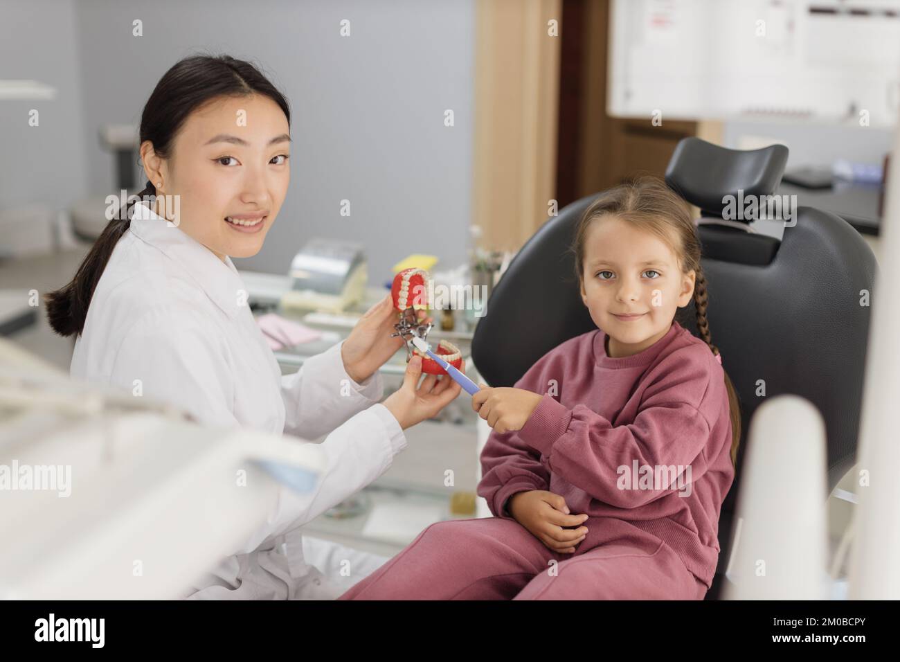 Happy smiling asian female dentist tells little child girl how to brush ...