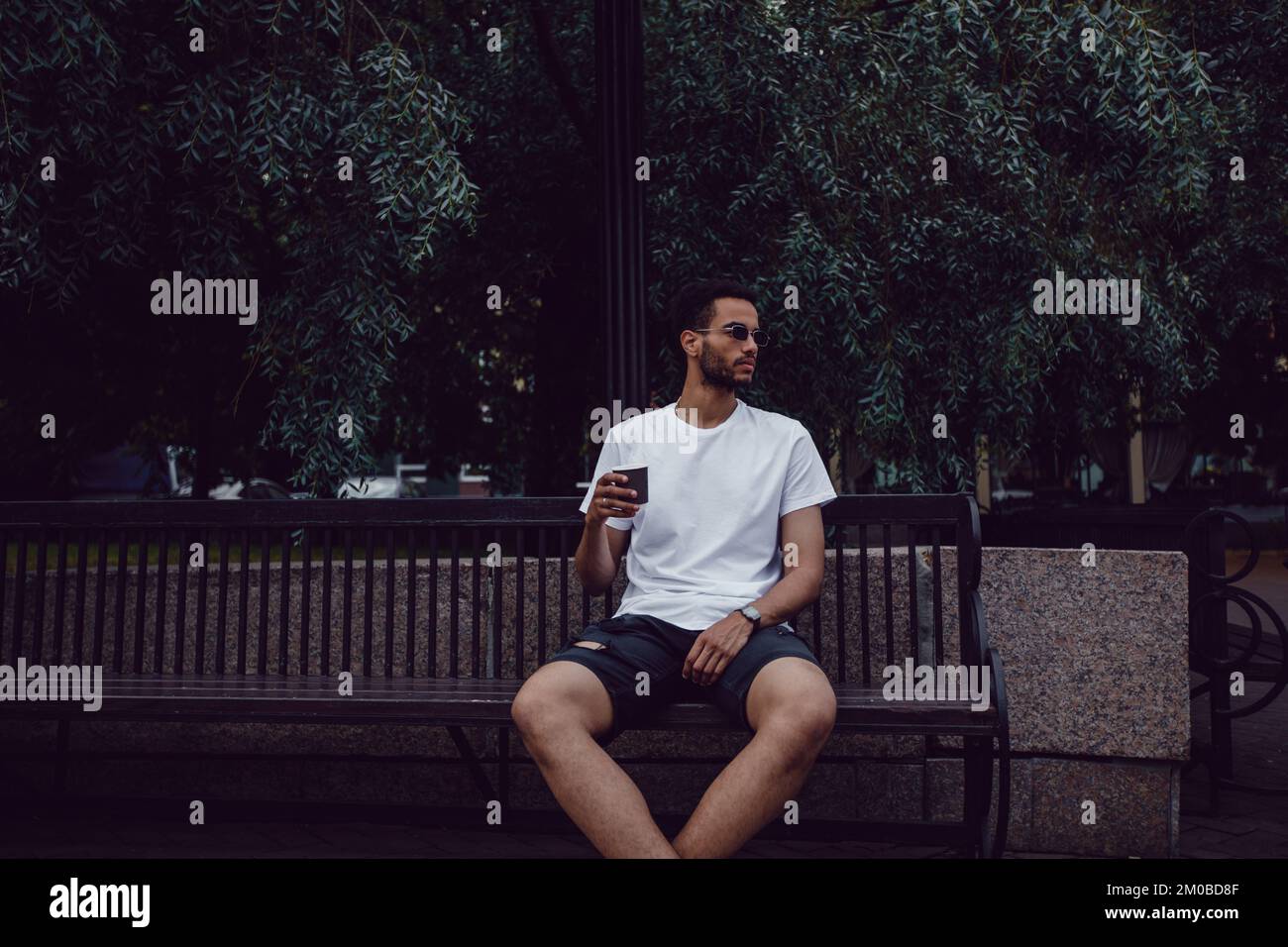 African american man in a white t-shirt sits on a bench and drinks ...