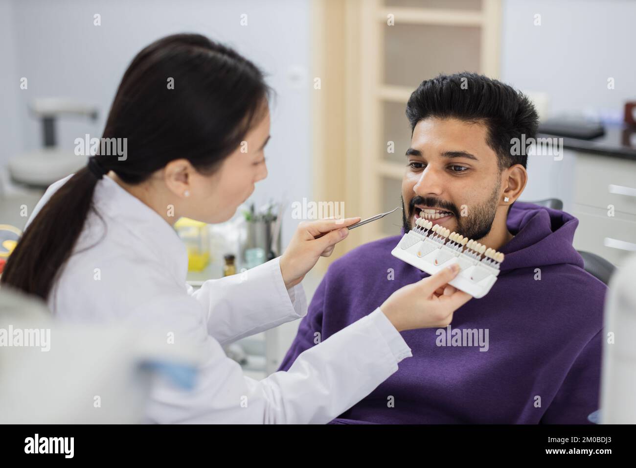 Asian dentist checking and selecting color of young bearded man teeth ...