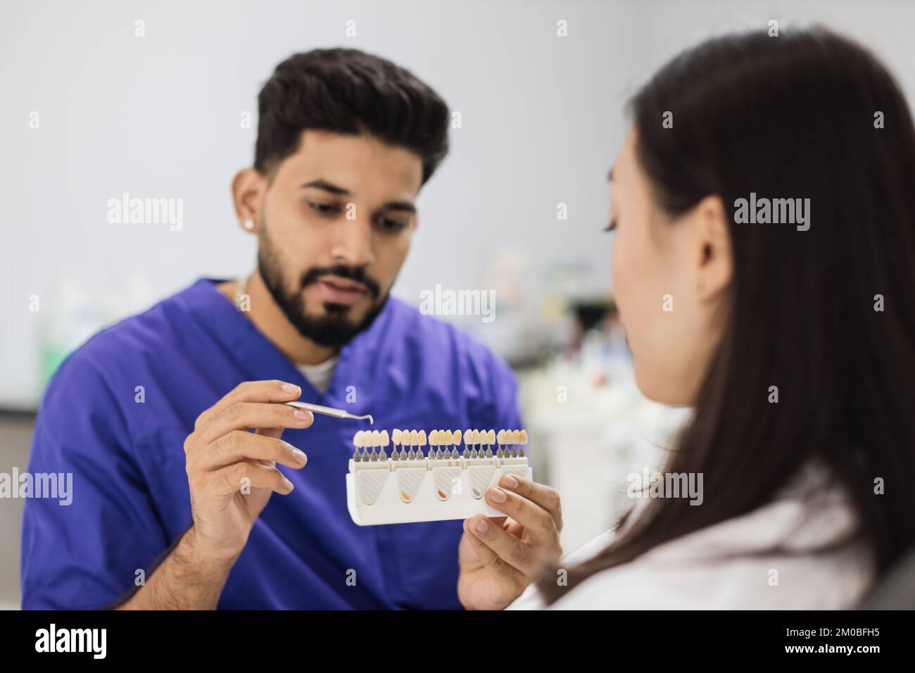 Bearded confident male dentist checking and selecting color of young woman teeth indoors in ...