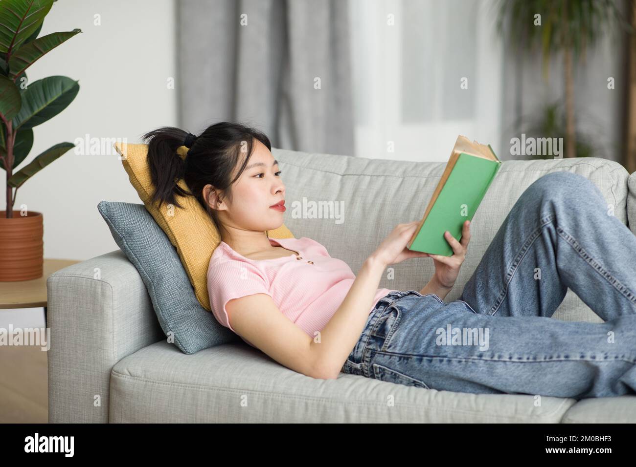 Carefree korean lady lying on couch in living room, reading book while ...
