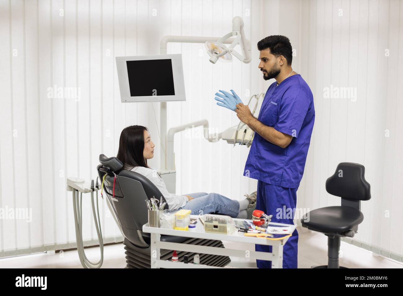 Confident bearded male dentist puts on blue rubber gloves, standing in a dental office, ready ...