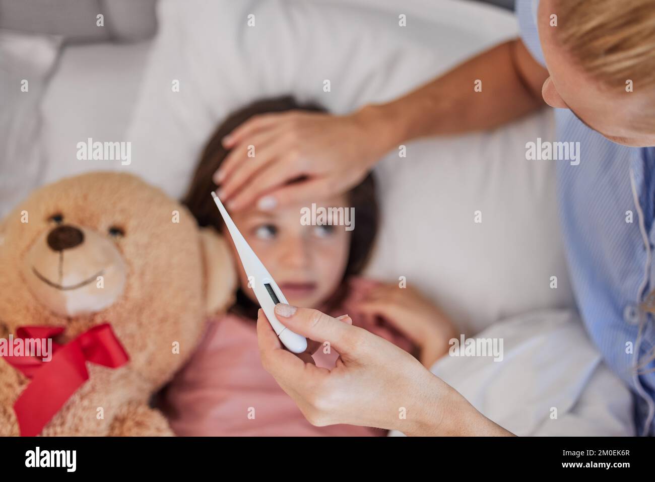 Sick little girl in bed with her teddybear while her mother uses a thermometer to check her ...
