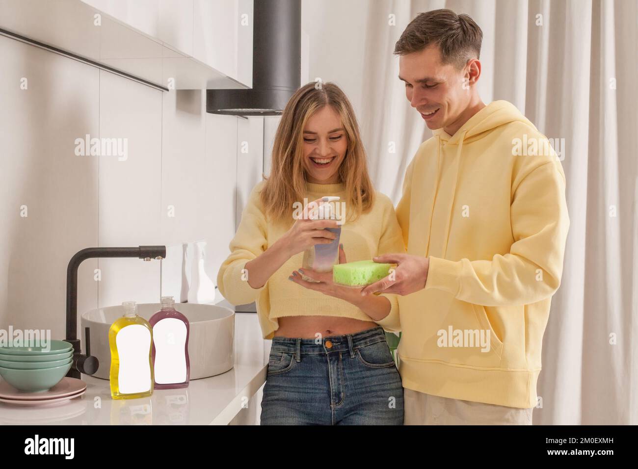 Joyful beautiful couple in yellow clothes holding bottle of dishwashing ...