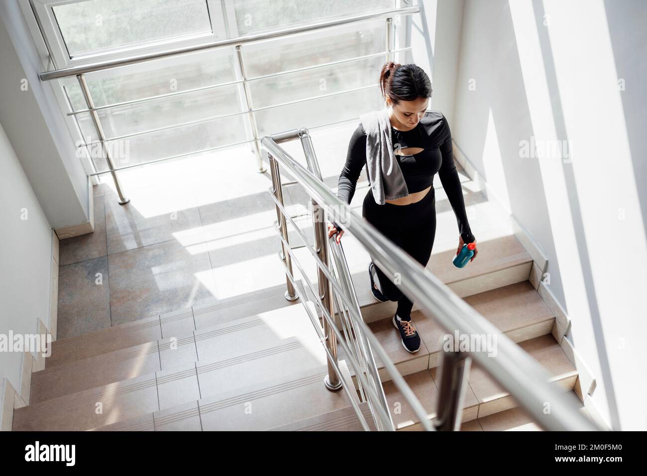 Cheerful sporty brunette woman with drink walking on stairs in gym in ...
