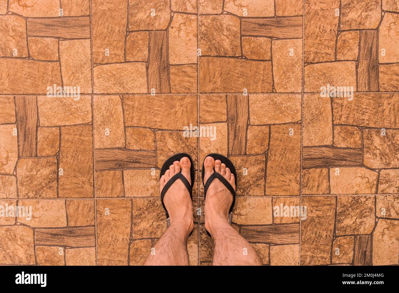 Legs of a man in flip flops stand on a brown tiled floor with an ...