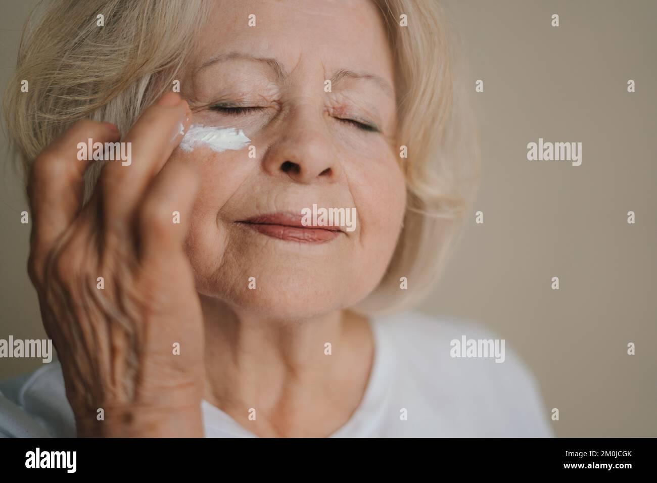 Senior woman with eyes closed putting tightening facial cream on face ...