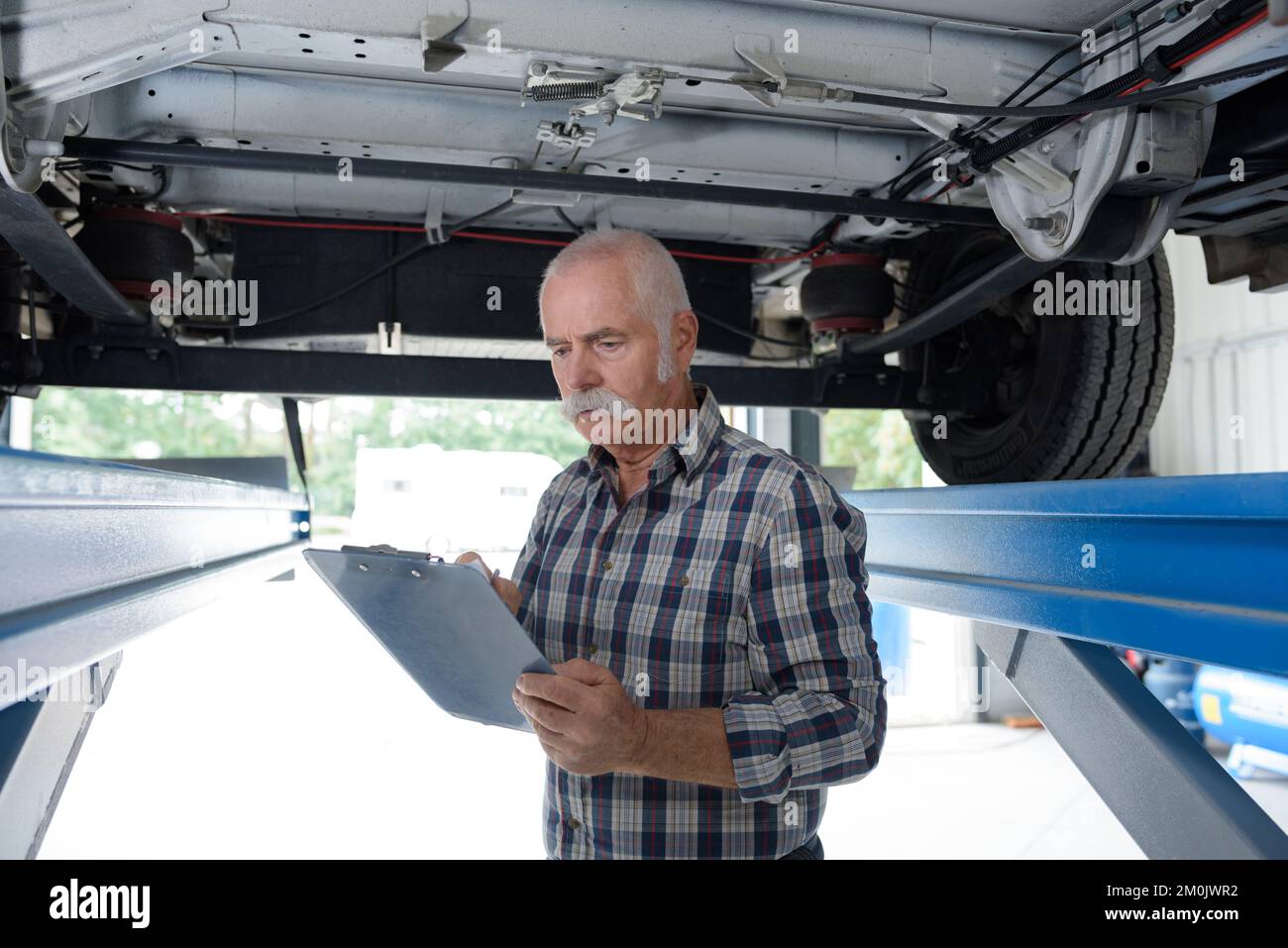 senior mechanic with clipboard working underneath elevated vehicle ...