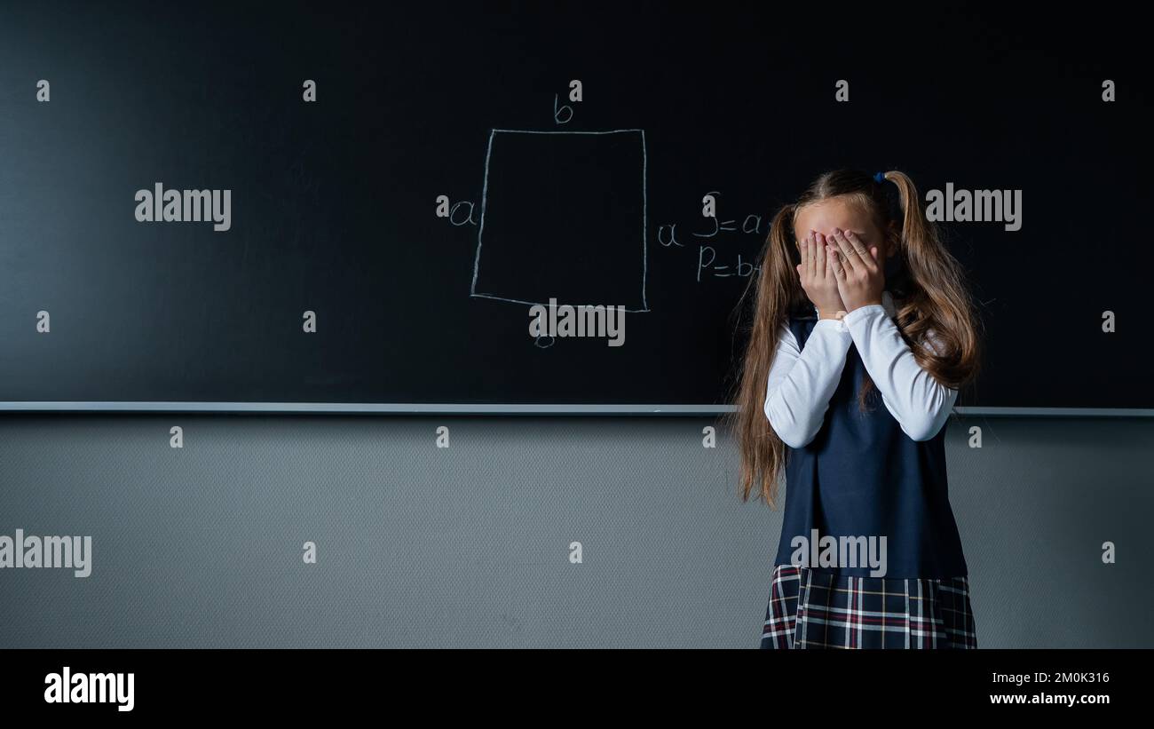 Caucasian girl stands at the blackboard and cries covering her face ...