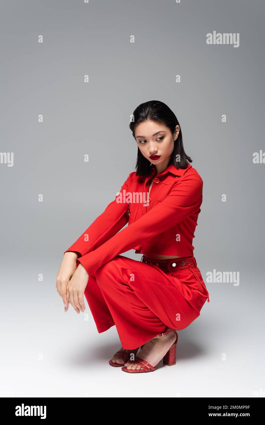 pretty asian woman in red stylish outfit sitting on haunches on grey ...