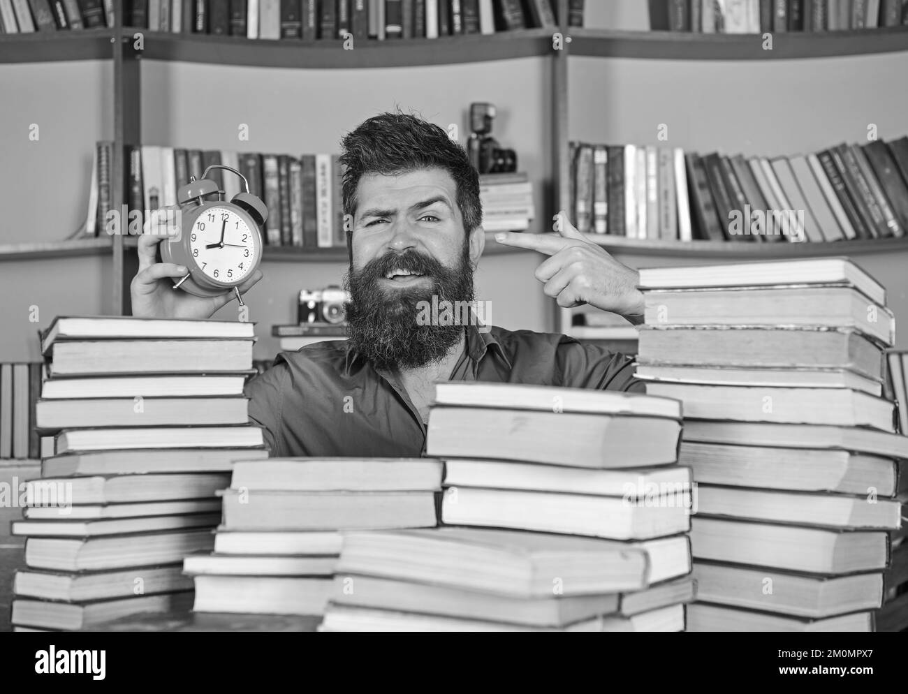 Man, scientist peeking out of piles of books with alarm clock. Teacher or student with beard ...