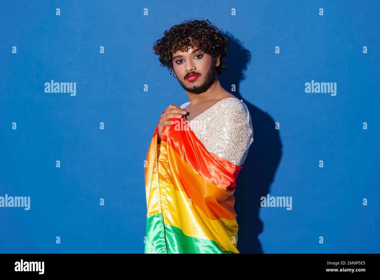 Young curly man wrapped rainbow flag looking at camera isolated over ...