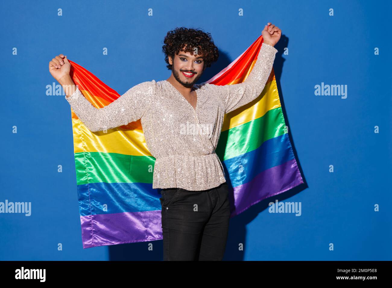 Young curly man with rainbow flag smiling at camera isolated over blue ...