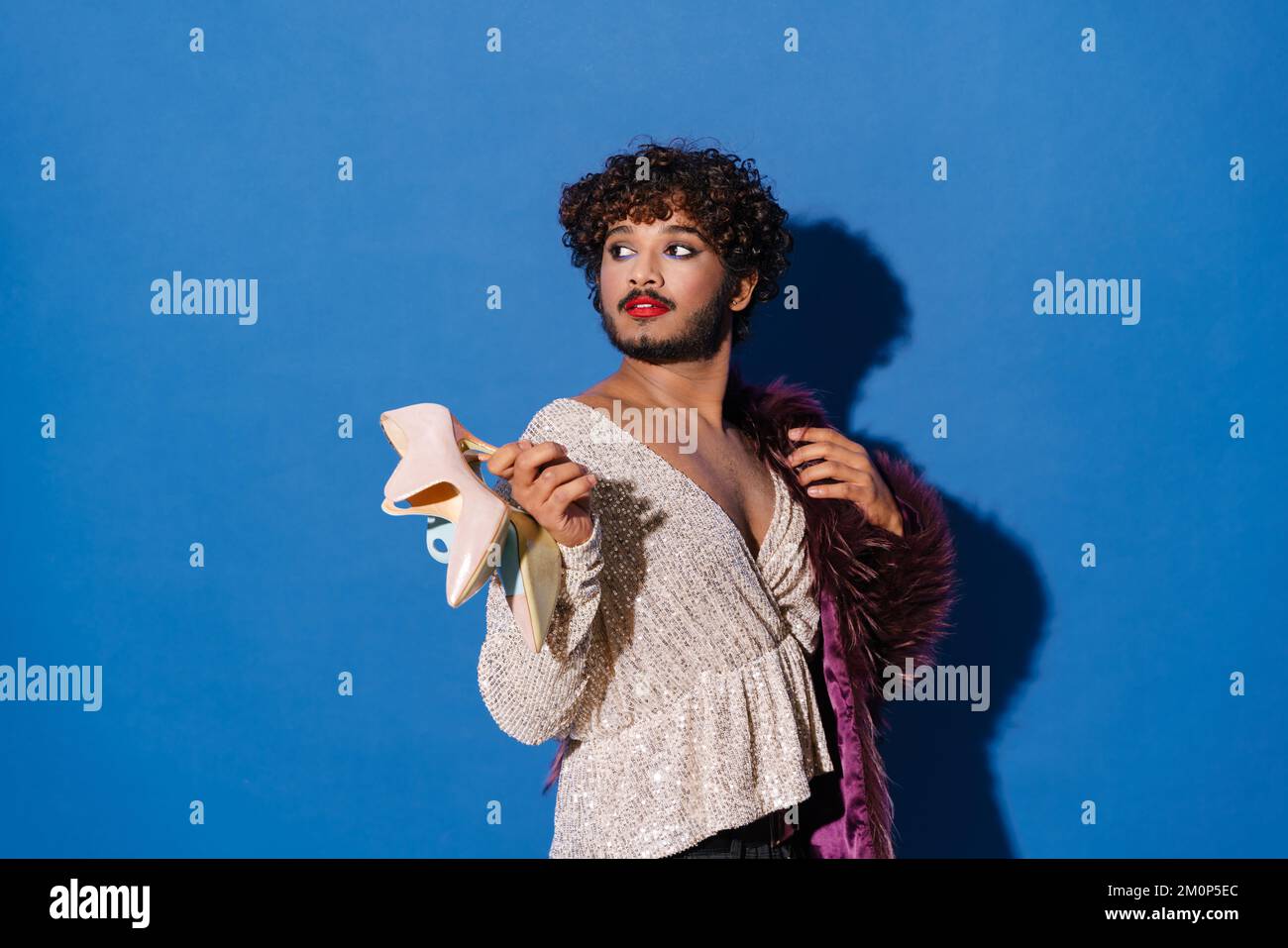 Young curly man with makeup holding high heels and looking aside ...