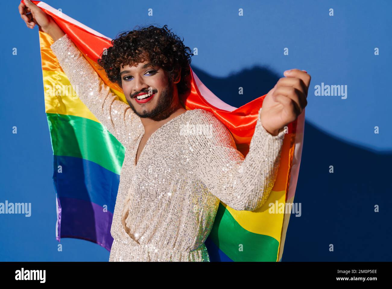 Young curly man with rainbow flag smiling at camera isolated over blue ...