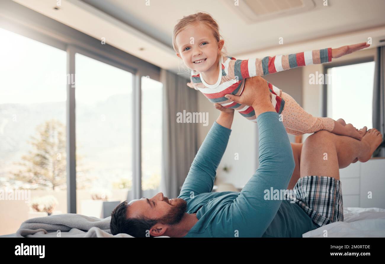 Adorable little girl being lifted in the air by her dad. Cheerful dad lying on bed and playing ...