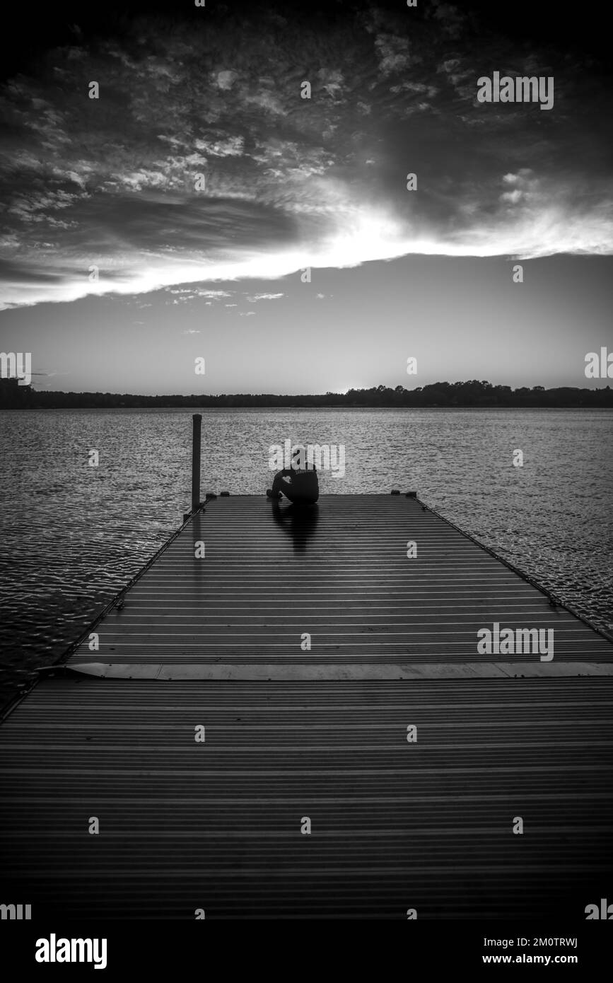 A vertical shot of a lonely fisherman sitting on a pier at a lake with ...
