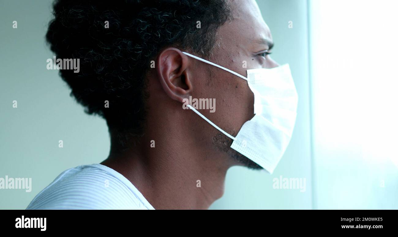 Black man in lockdown staying at home looking out window wearing mask ...