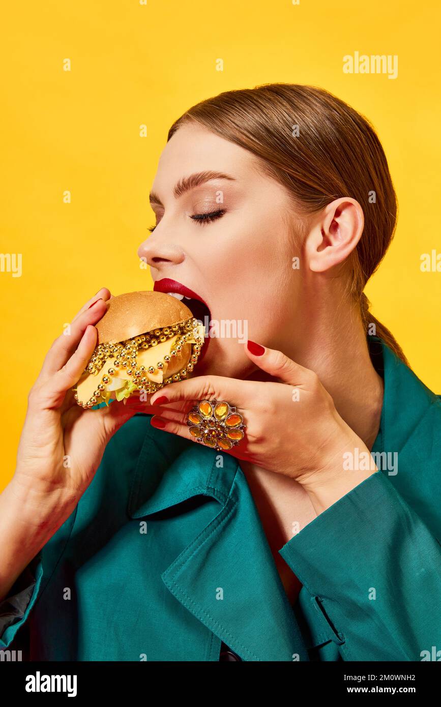 Young beautiful woman in green coat eating, biting cheeseburger with ...