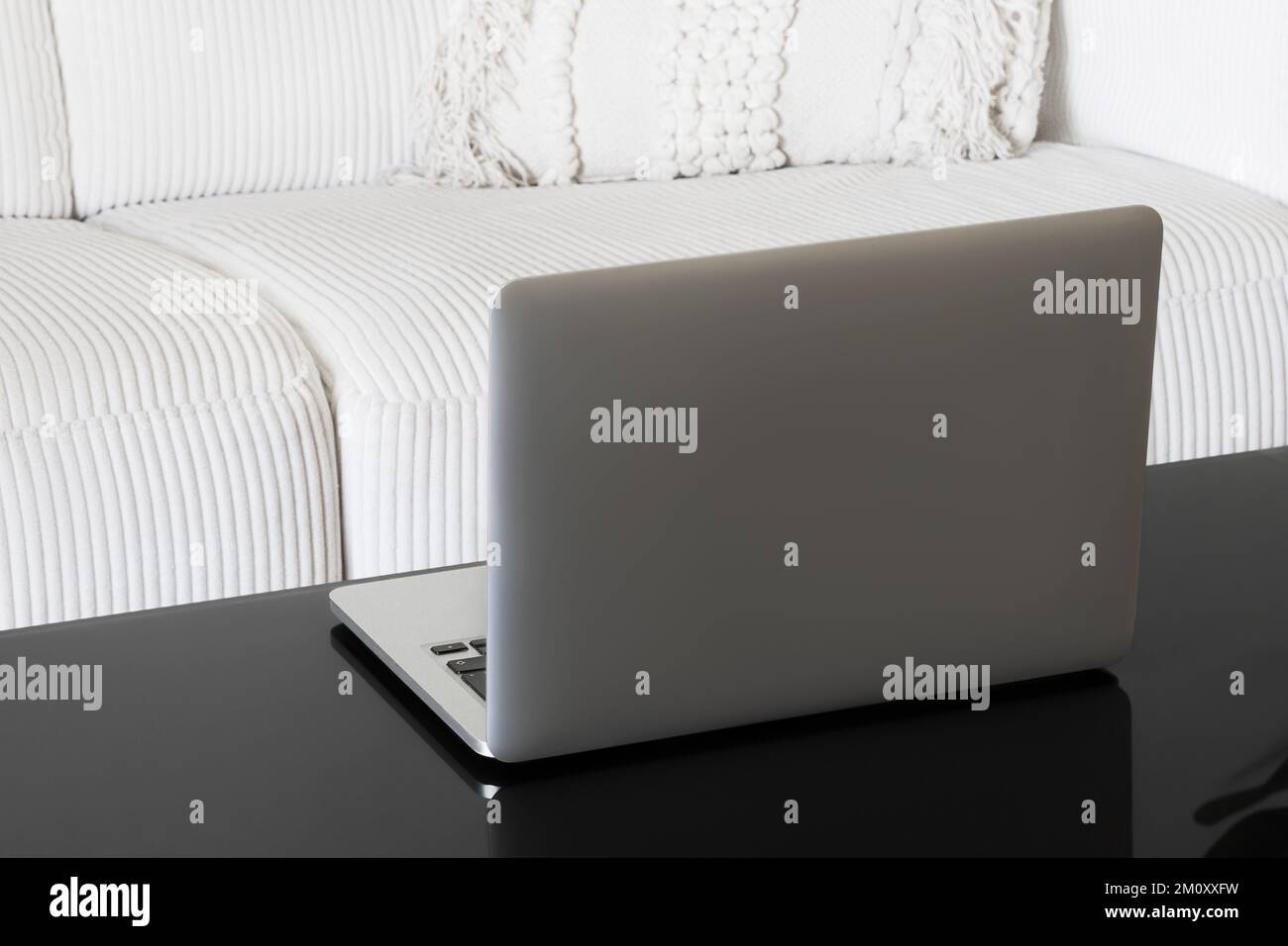 Laptop on a black glass table against a white sofa. Back view. The ...