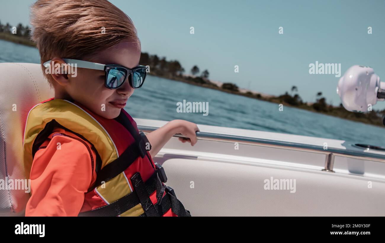 A blond boy wearing a life jacket and sunglasses sitting in a boat ...