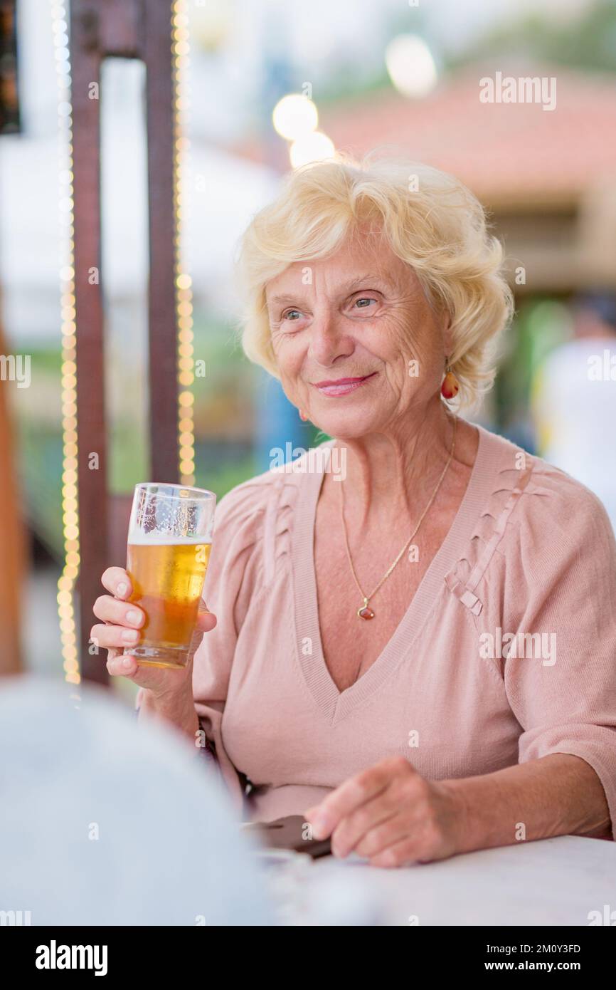 Mature woman sitting at a table in a summer cafe and drinking beer ...
