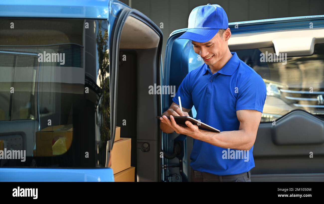 Smiling asian male courier in blue uniform using digital tablet next to ...