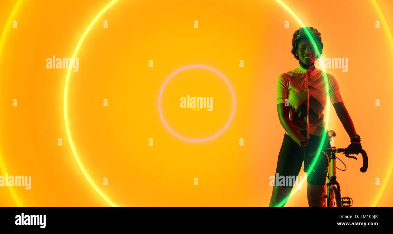 Biracial smiling female athlete with bike standing by illuminated circles on yellow background ...