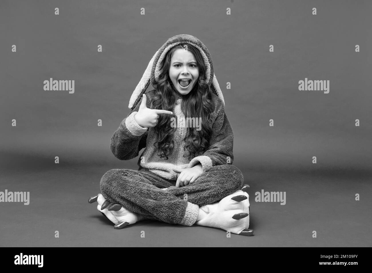 amazed kid in comfortable pajama sit pointing finger on herself on red ...