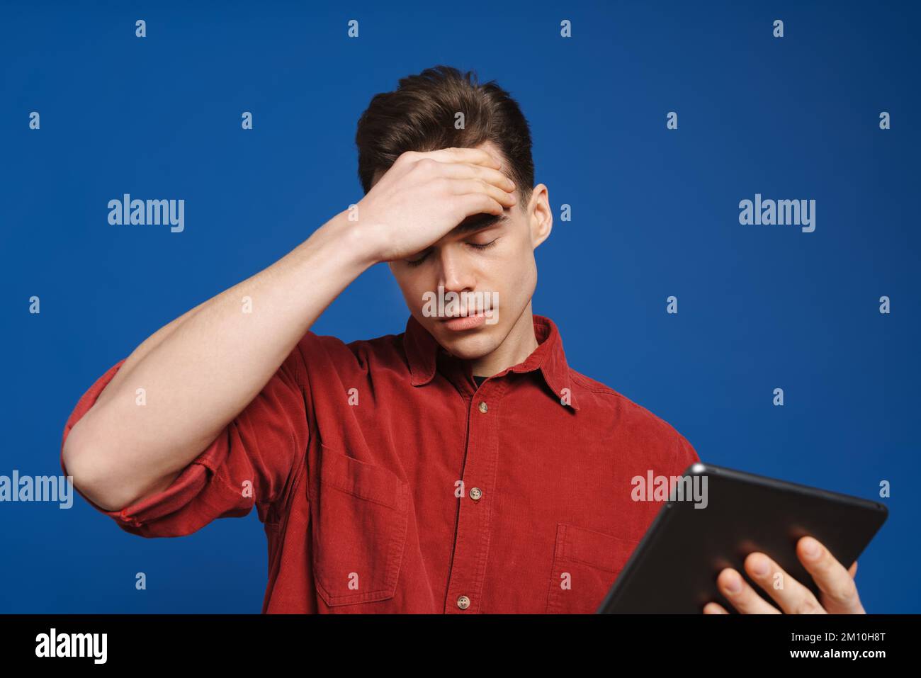 Young white man with headache using tablet computer isolated over blue ...