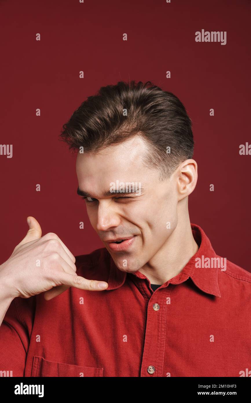 Young white man making handset gesture and winking isolated over red ...