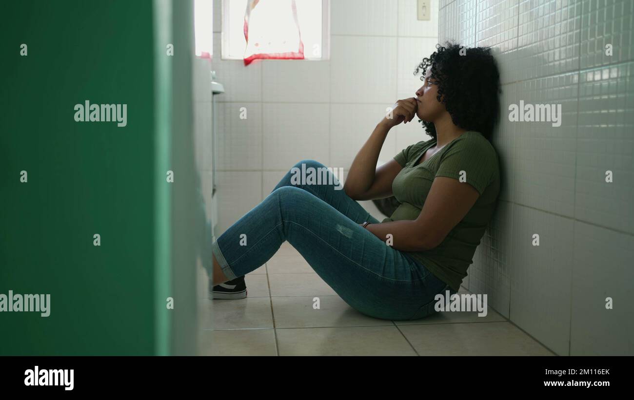 A preoccupied young black woman seated on kitchen floor feeling lonely ...