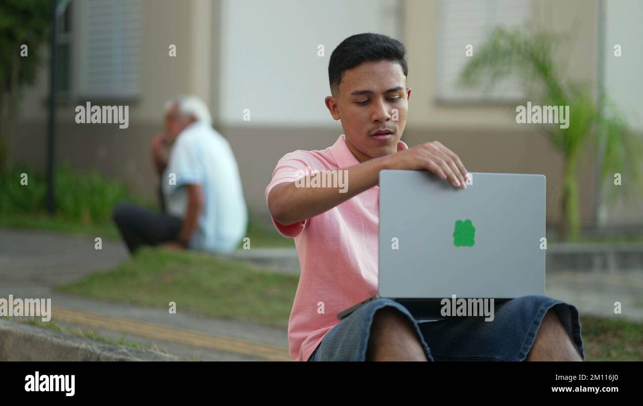 A young hispanic man opening laptop computer screen outside. Candid ...