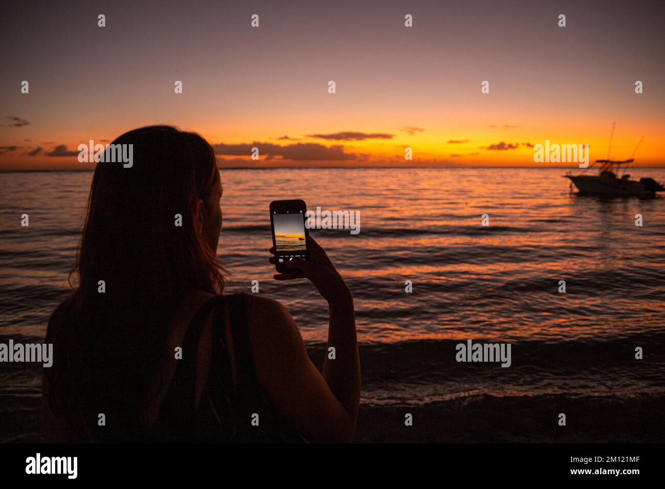A young lady taking a picture of the sunset at a beach in Mauritius ...