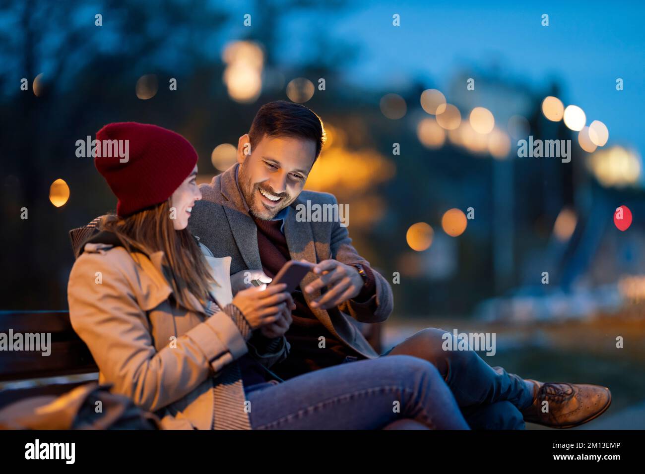 Happy young couple sitting in a park bench and using smart phone ...