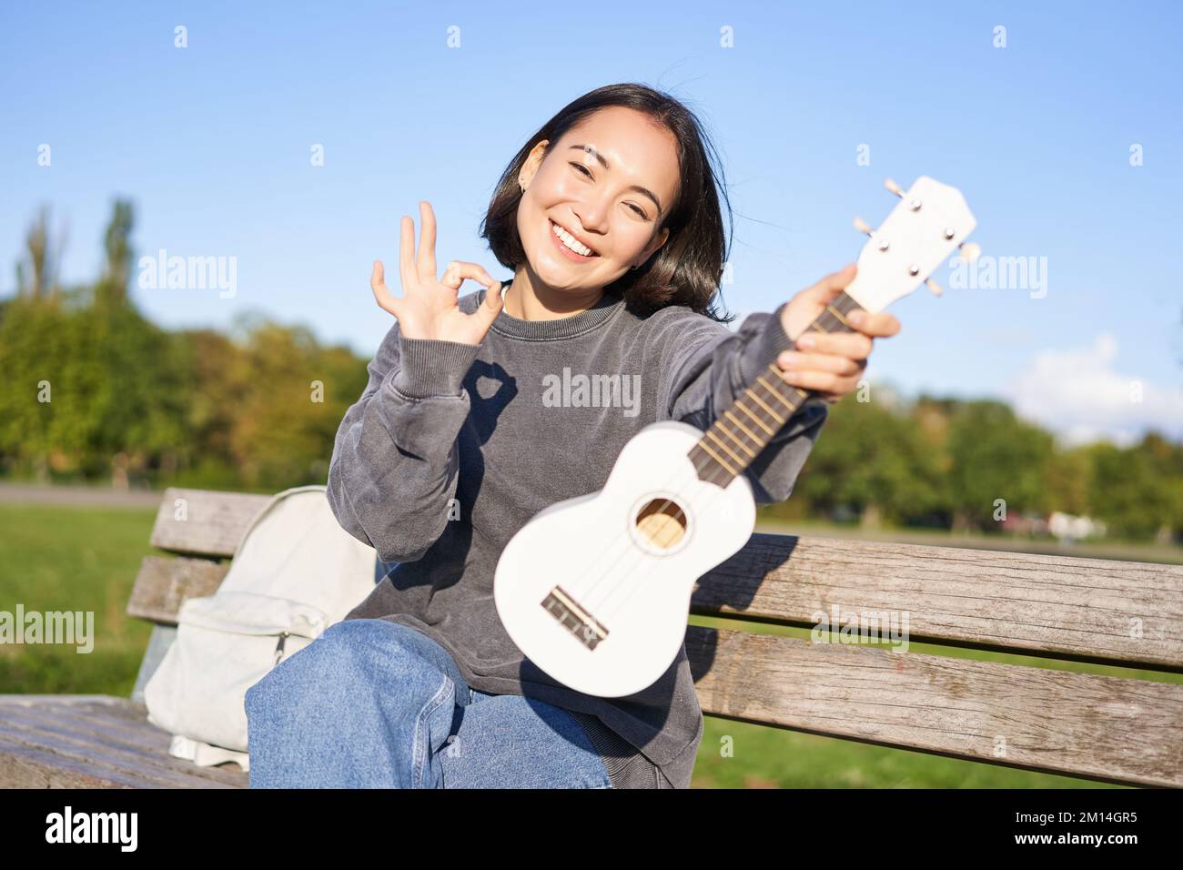 Cute smiling girl shows ok sign and her new ukulele, sits on bench in ...