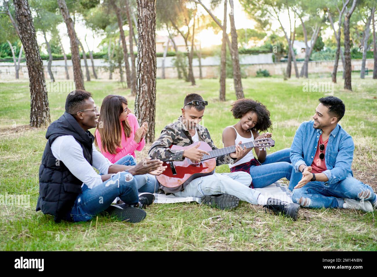 Multi-ethnic friends sitting on the ground while one plays guitar in a ...