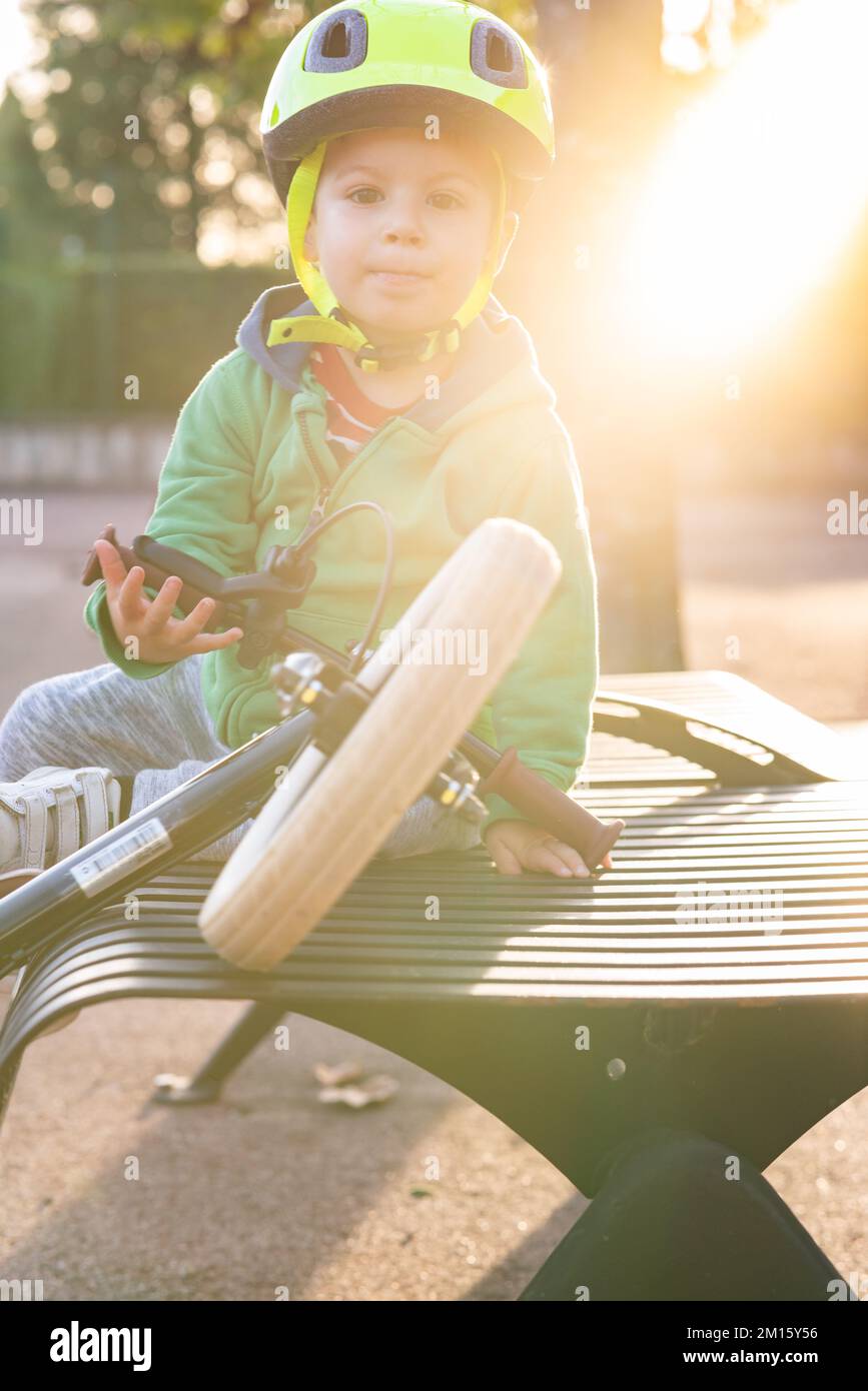 Little boy in yellow helmet resting on bench near bike and looking at ...