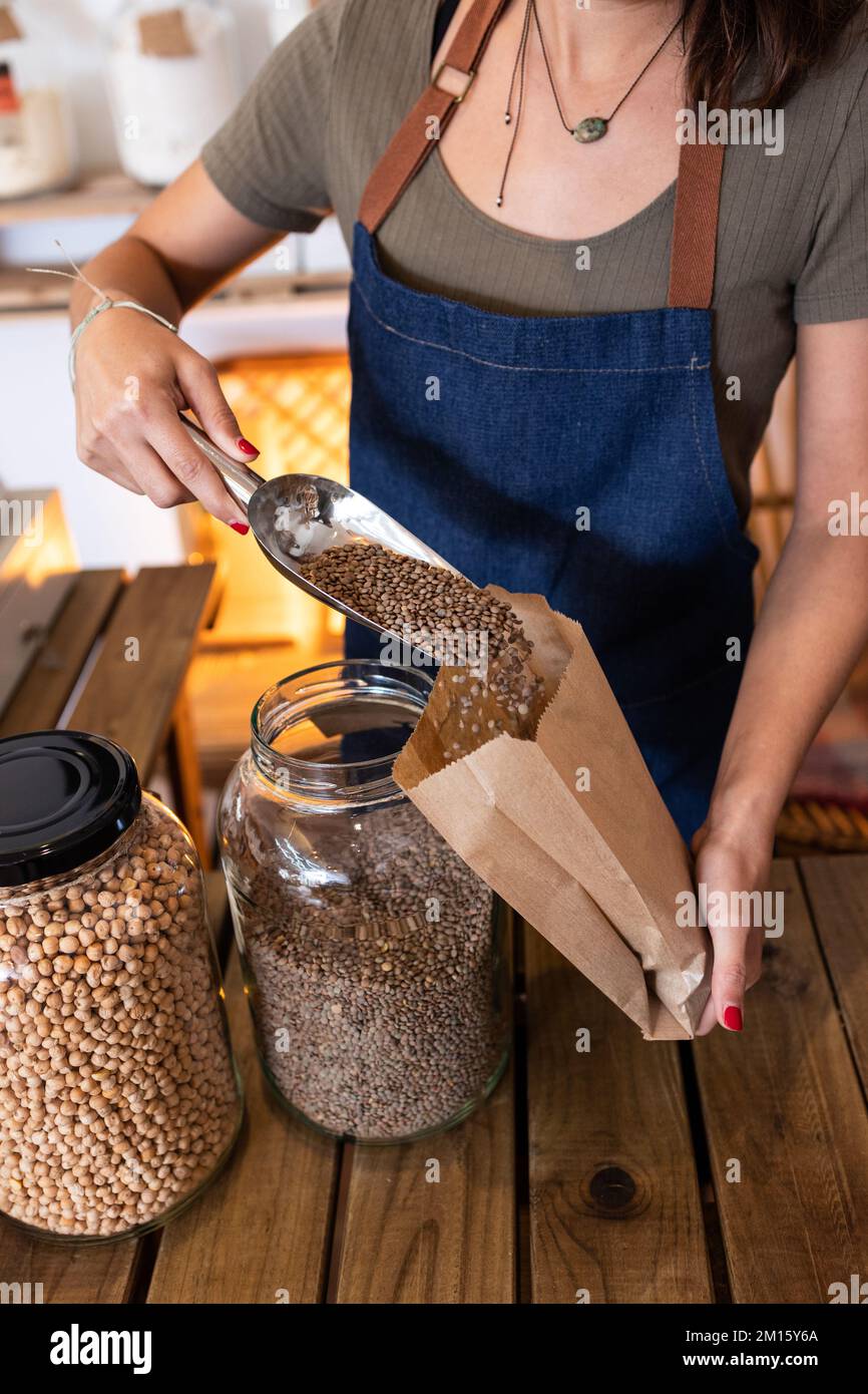Crop unrecognizable female in apron standing at counter with scoop while pouring brown seeds ...